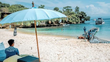 Uma praia com um guarda-sol azul, uma placa, uma pessoa com prancha de surf e barcos na água.