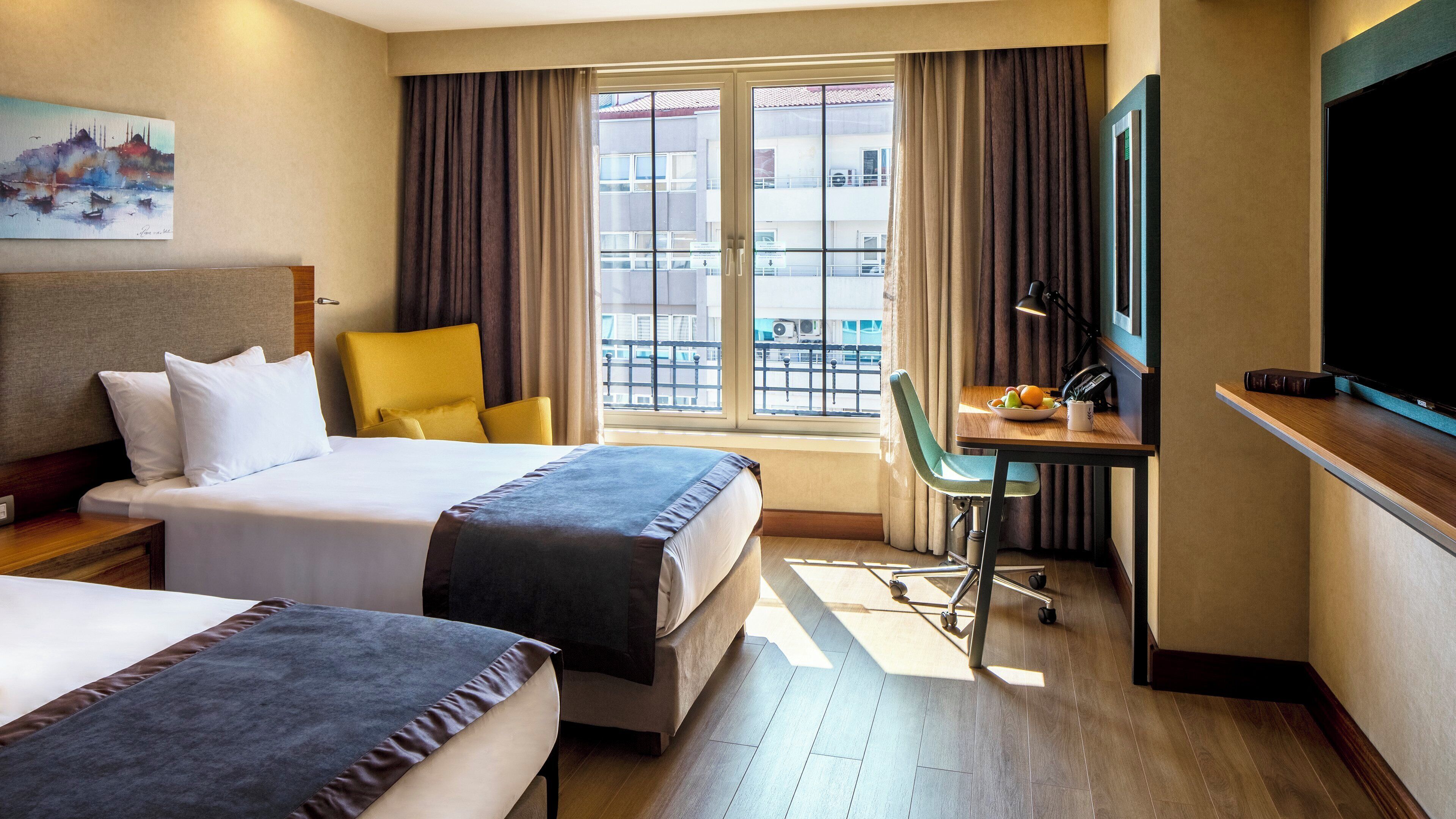 Quarto de hotel com duas camas, uma mesa de trabalho com uma cadeira, televisão e janela com cortinas.