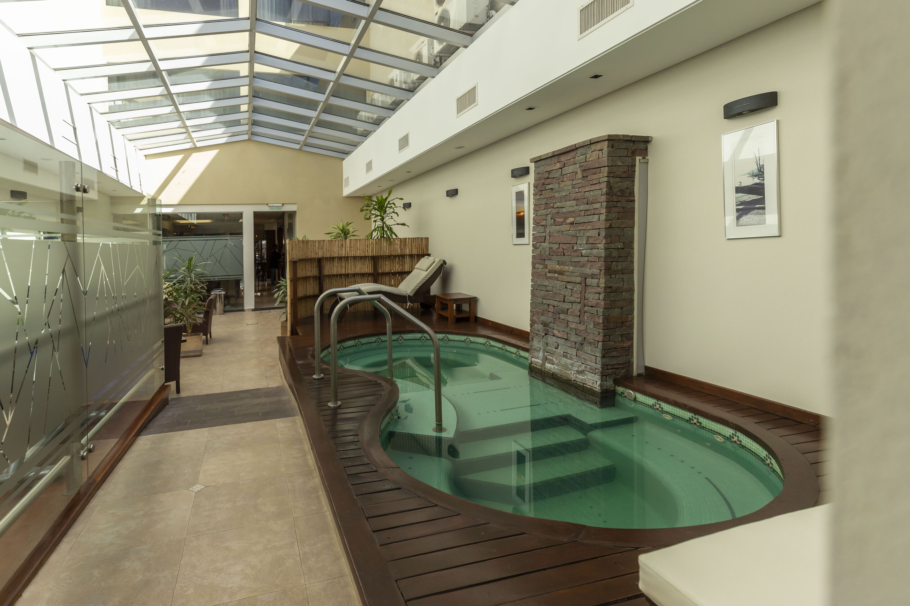 Piscina interna moderna com teto de vidro, deck de madeira e parede de pedra.