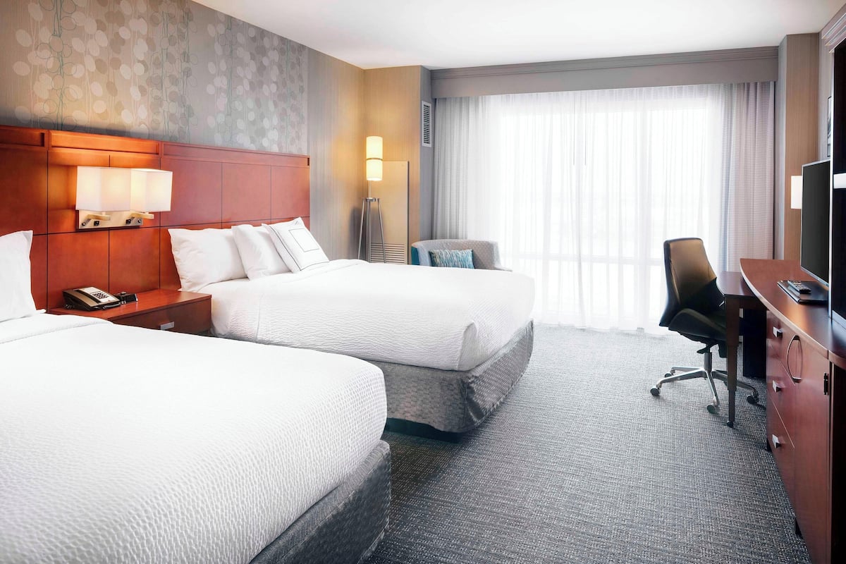 Quarto de hotel com duas camas, uma escrivaninha, uma cadeira, um abajur e uma televisão.