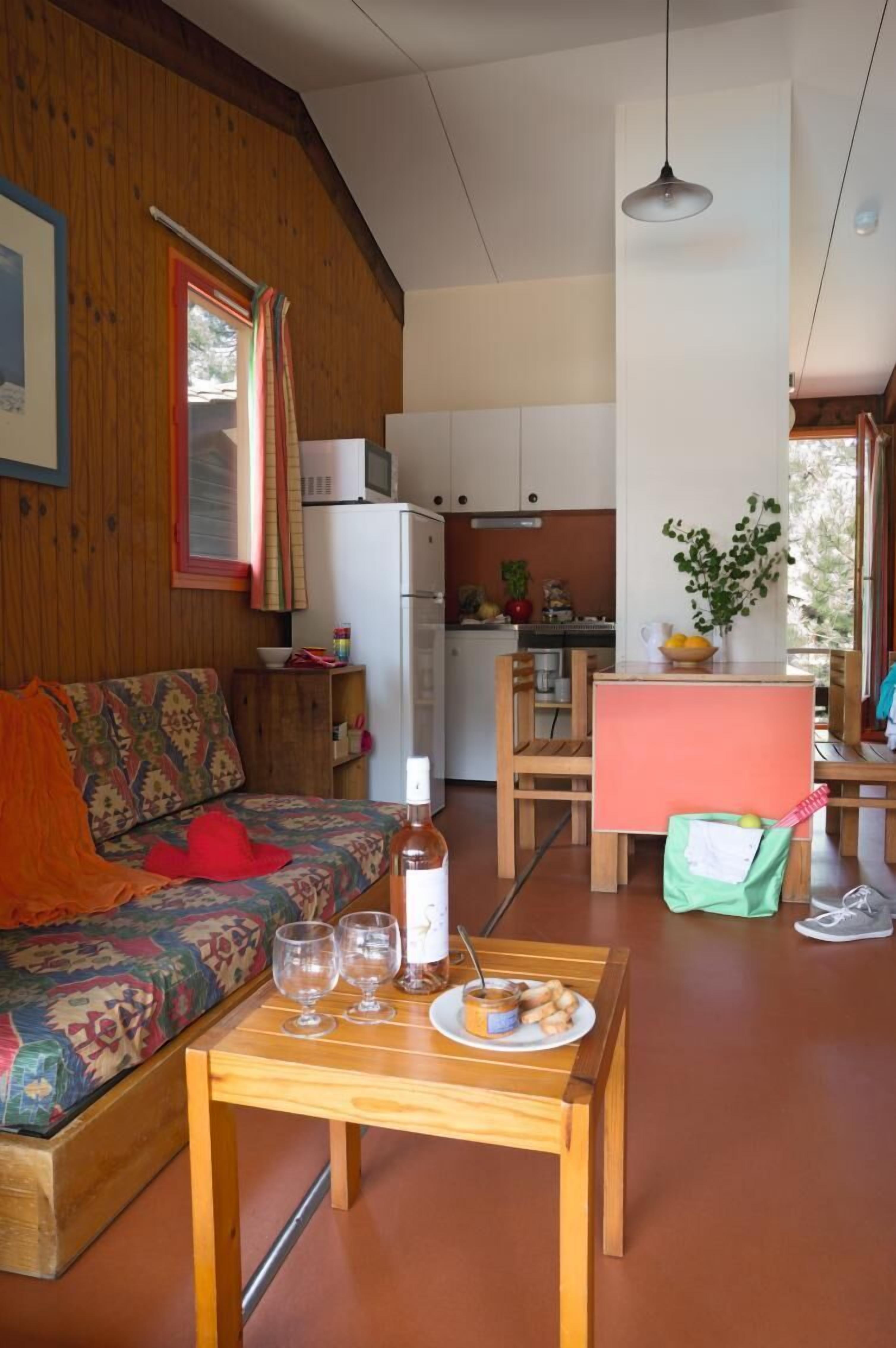 Uma sala de estar aconchegante com um sofá, uma mesa de madeira com um prato de comida e copos, uma área de cozinha com geladeira e micro-ondas, e uma sala de jantar com mesa e cadeiras.
