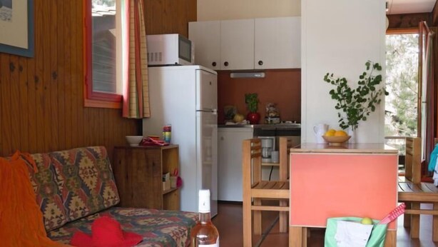 Uma sala de estar aconchegante com um sofá, uma mesa de madeira com um prato de comida e copos, uma área de cozinha com geladeira e micro-ondas, e uma sala de jantar com mesa e cadeiras.