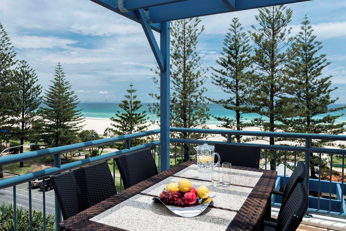 Uma mesa com uma cesta de frutas, jarra e copos, com vista para uma praia e árvores.