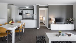 Uma sala de estar moderna com um sofá branco, uma mesa de centro e uma televisão. Há uma área de jantar com mesa e cadeiras brancas.