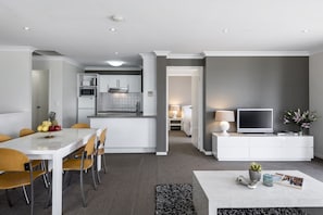 Uma sala de estar moderna com um sofá branco, uma mesa de centro e uma televisão. Há uma área de jantar com mesa e cadeiras brancas.