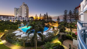 Um resort com piscina rodeada por palmeiras e espreguiçadeiras, com vista para um corpo de água e edifícios altos.