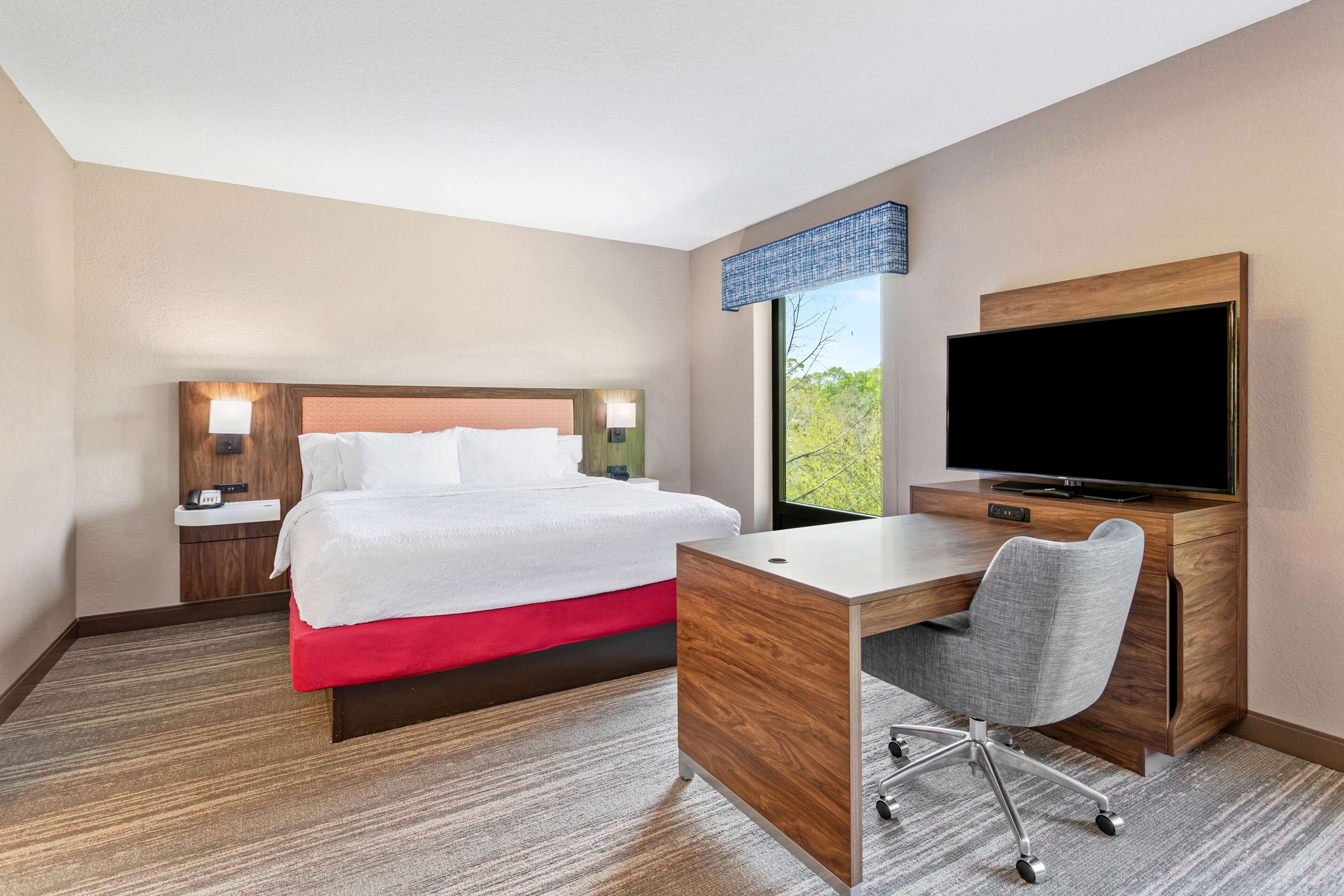 Quarto de hotel com uma cama, uma escrivaninha, uma cadeira, uma televisão e uma janela com vista para árvores.