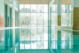 Uma grande piscina interior com paredes de vidro transparente, chão de azulejo e vista para árvores lá fora.