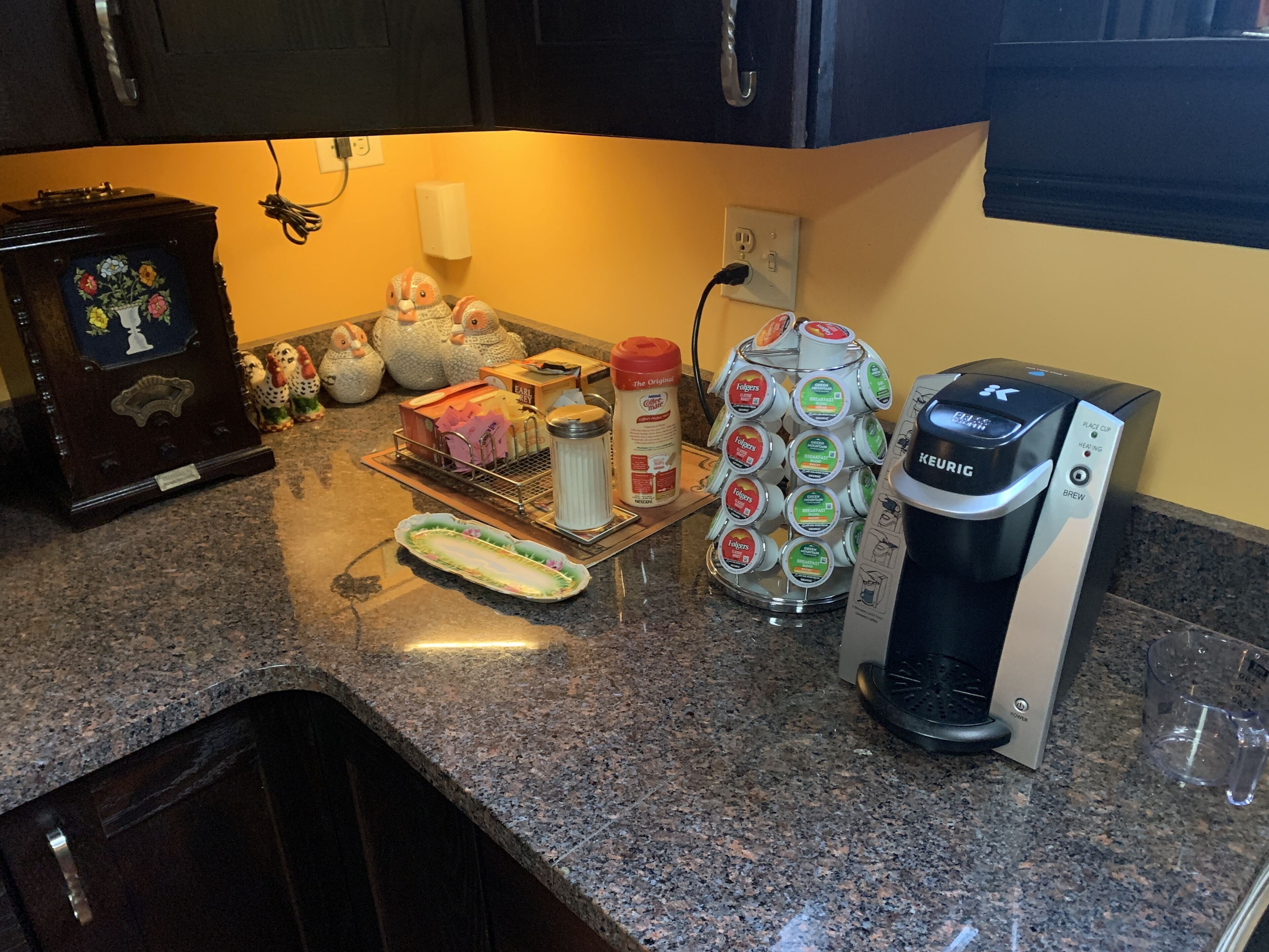Uma bancada de cozinha com uma cafeteira, uma chaleira, um tabuleiro com saquinhos de chá e um porquinho de cerâmica decorativo.