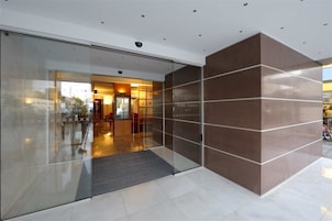 Entrada moderna de hotel com porta de vidro, área de recepção com assentos e parede marrom com o nome do hotel.