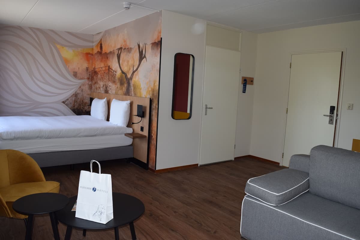Quarto de hotel com cama, sofá, mesa pequena, cadeira, pintura e espelho.