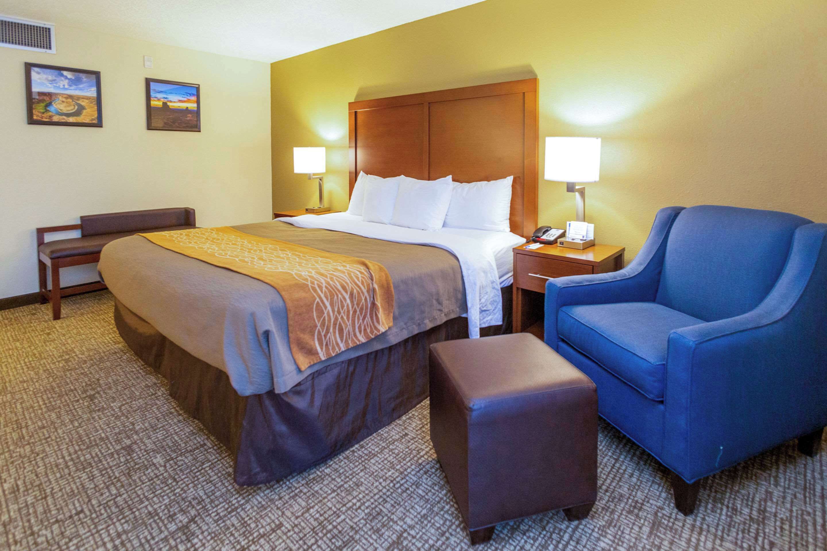 Quarto de hotel com uma cama grande, uma poltrona azul, uma mesa pequena e duas luminárias de cabeceira.
