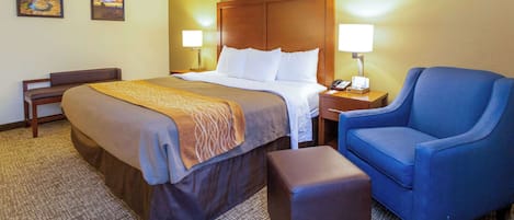 Quarto de hotel com uma cama grande, uma poltrona azul, uma mesa pequena e duas luminárias de cabeceira.