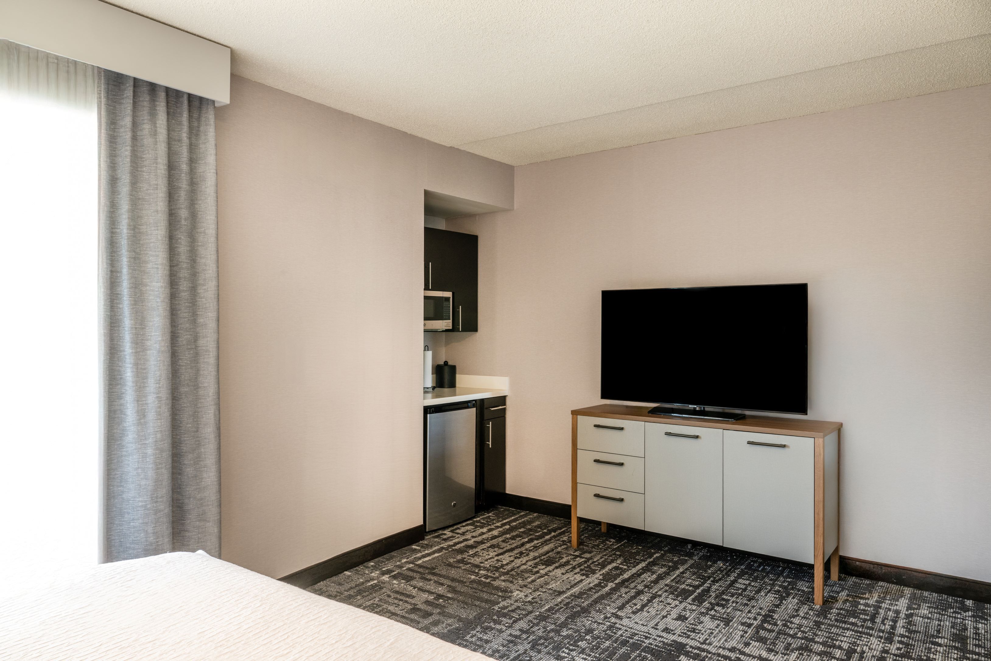 Quarto de hotel com cama, televisão de tela plana, pequeno espaço de cozinha e piso coberto de carpete.