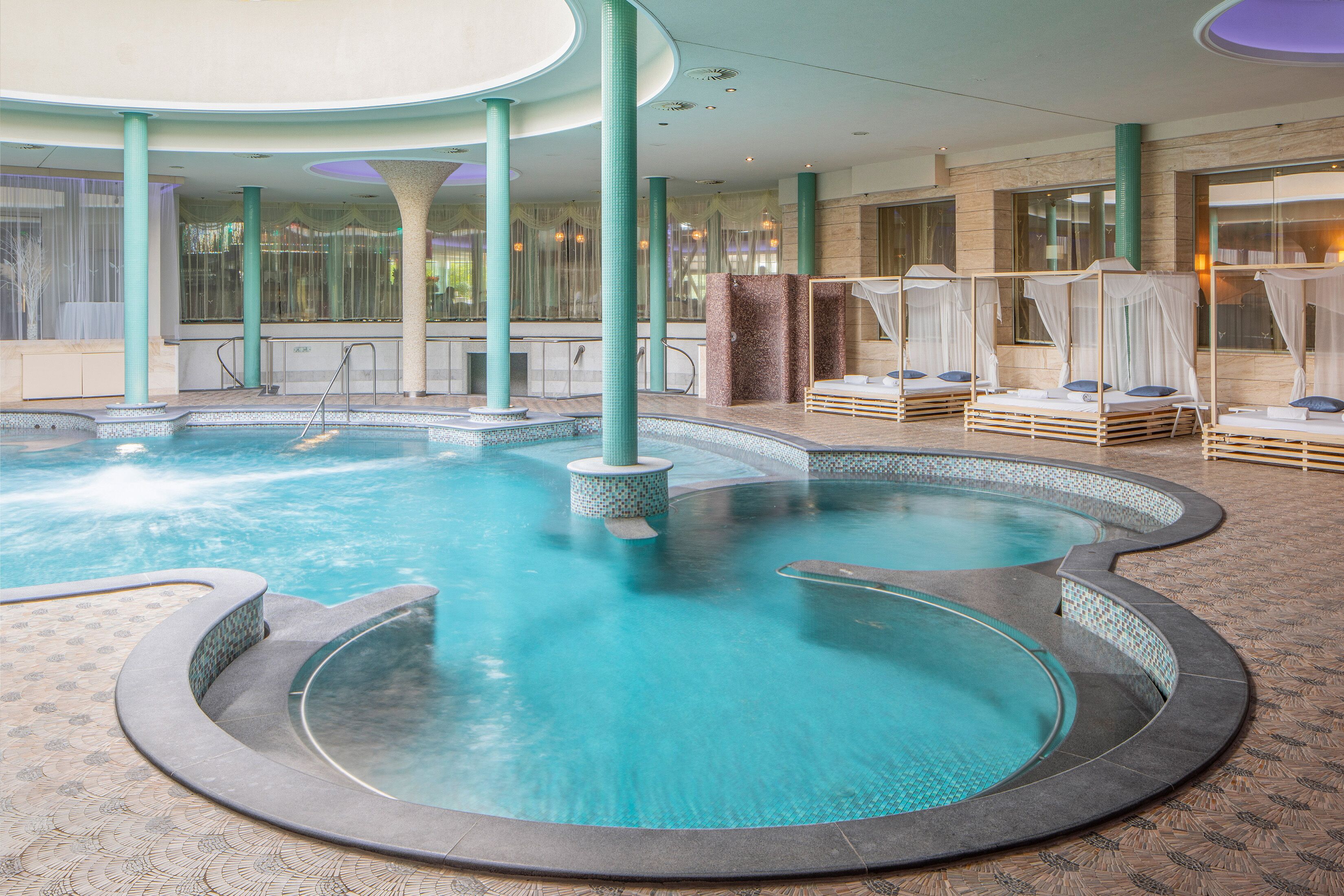 Piscina circular interior com uma fonte de água azul cristalina, rodeada por espreguiçadeiras e um chão em mosaico.