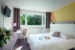 Quarto de hotel com cama, escrivaninha, cadeira, televisão, janela com cortinas e mesa de cabeceira com bandeja de suco de laranja.