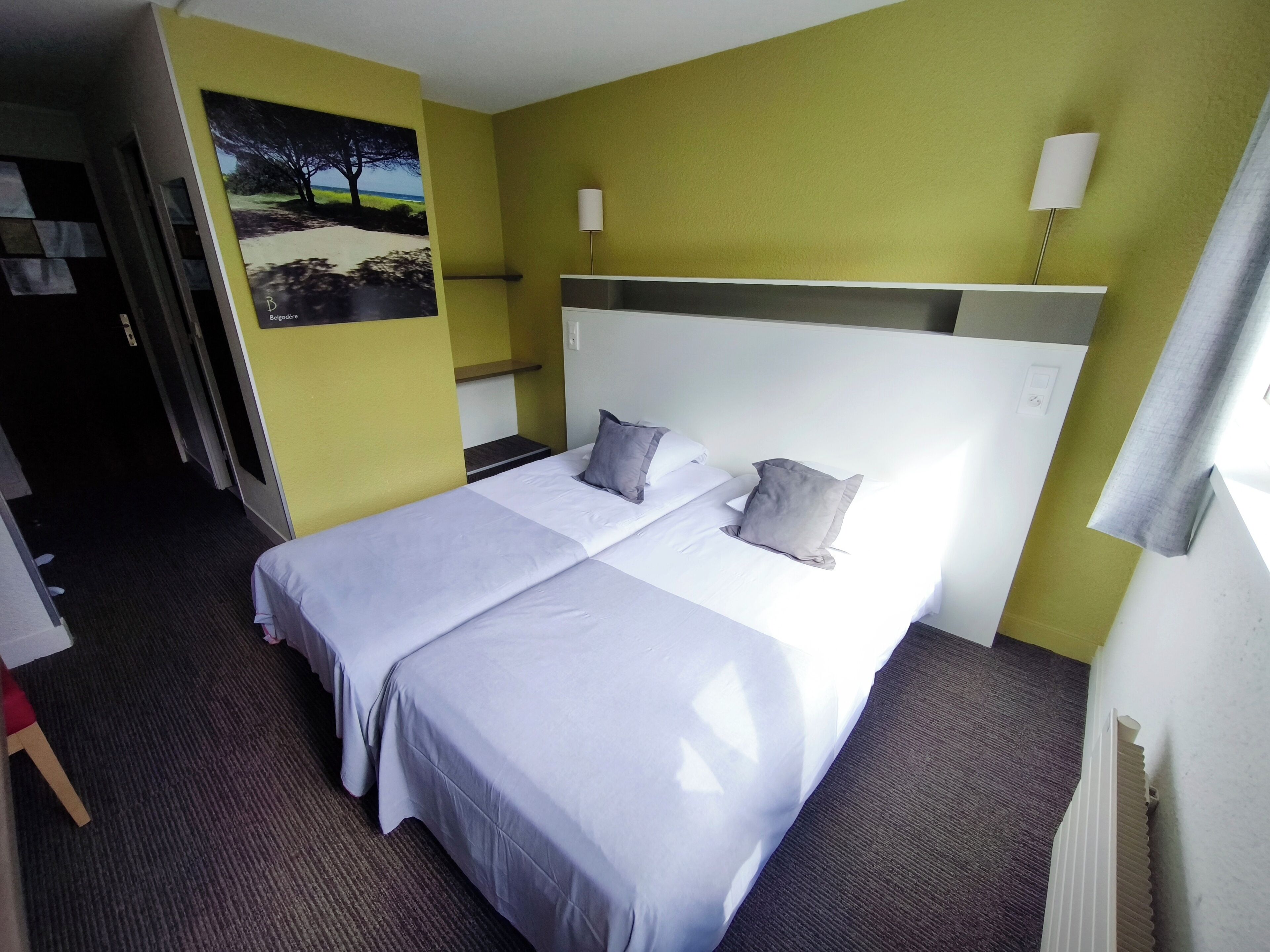 Quarto de hotel com uma cama grande, duas almofadas, mesa de cabeceira, abajur e um quadro na parede.
