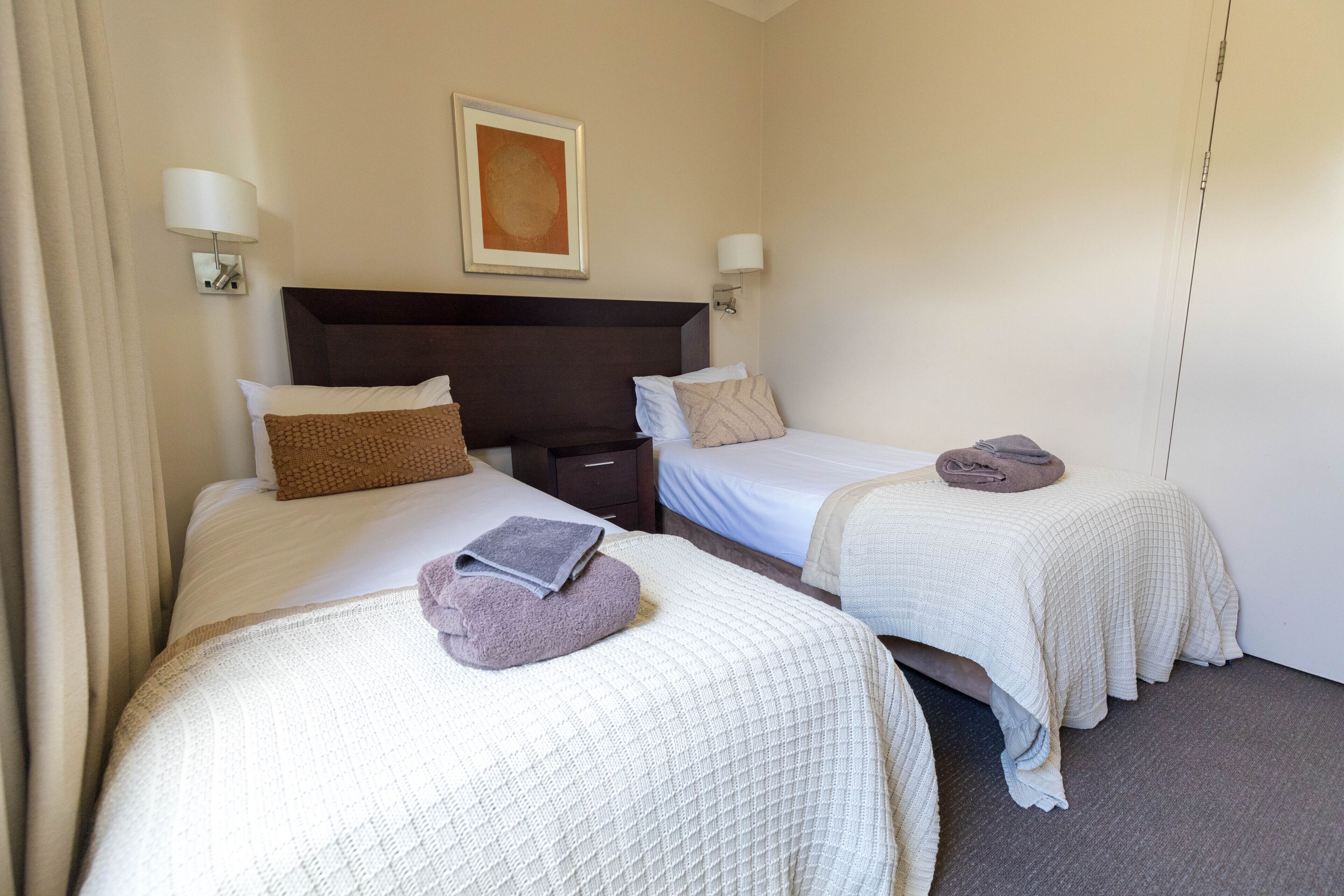 Quarto de hotel com duas camas, cada uma com almofada e toalha dobrada. Há mesas de cabeceira com luminárias e um quadro na parede acima das camas.