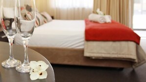 Quarto de hotel com cama, mesa de cabeceira com dois copos e uma flor, e janela com cortinas.