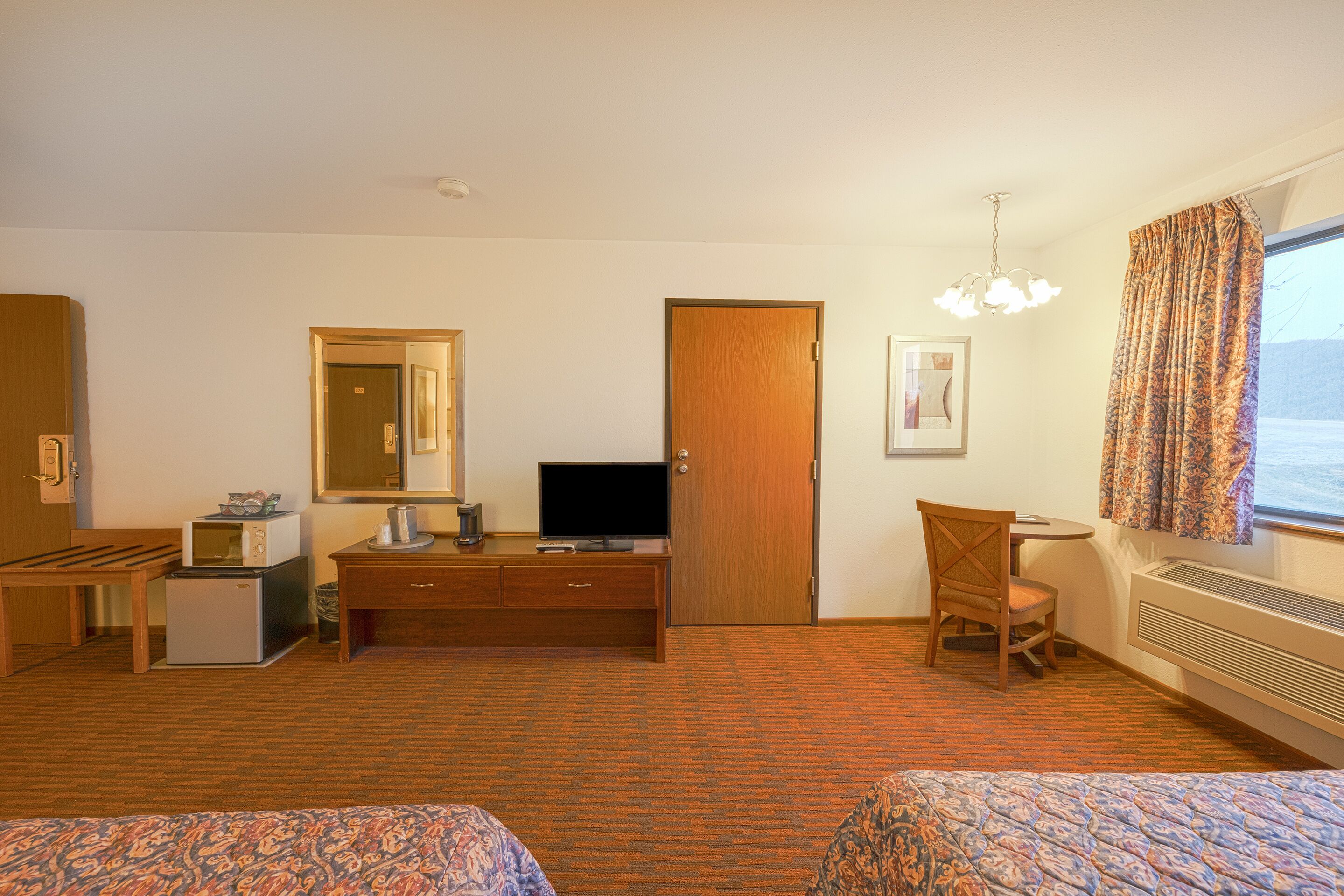 Quarto de hotel com duas camas, uma escrivaninha com computador, micro-ondas, mini-geladeira, televisão, uma cadeira e janela com cortinas.