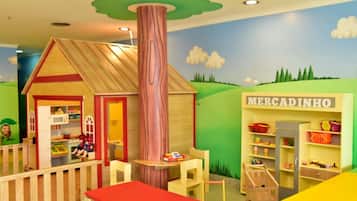 Uma sala de jogos infantil com uma casinha de madeira, um tronco de árvore grande e estantes com a etiqueta 'MERCADINHO'.