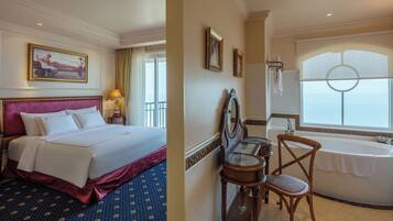 Quarto de hotel com uma cama grande, área de estar com poltrona e mesa, e banheiro separado com banheira.