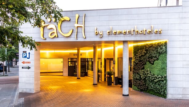 A entrada de um hotel chamado 'TACH by ClementHotels', com design moderno, incluindo uma parede verde e sinalização iluminada.