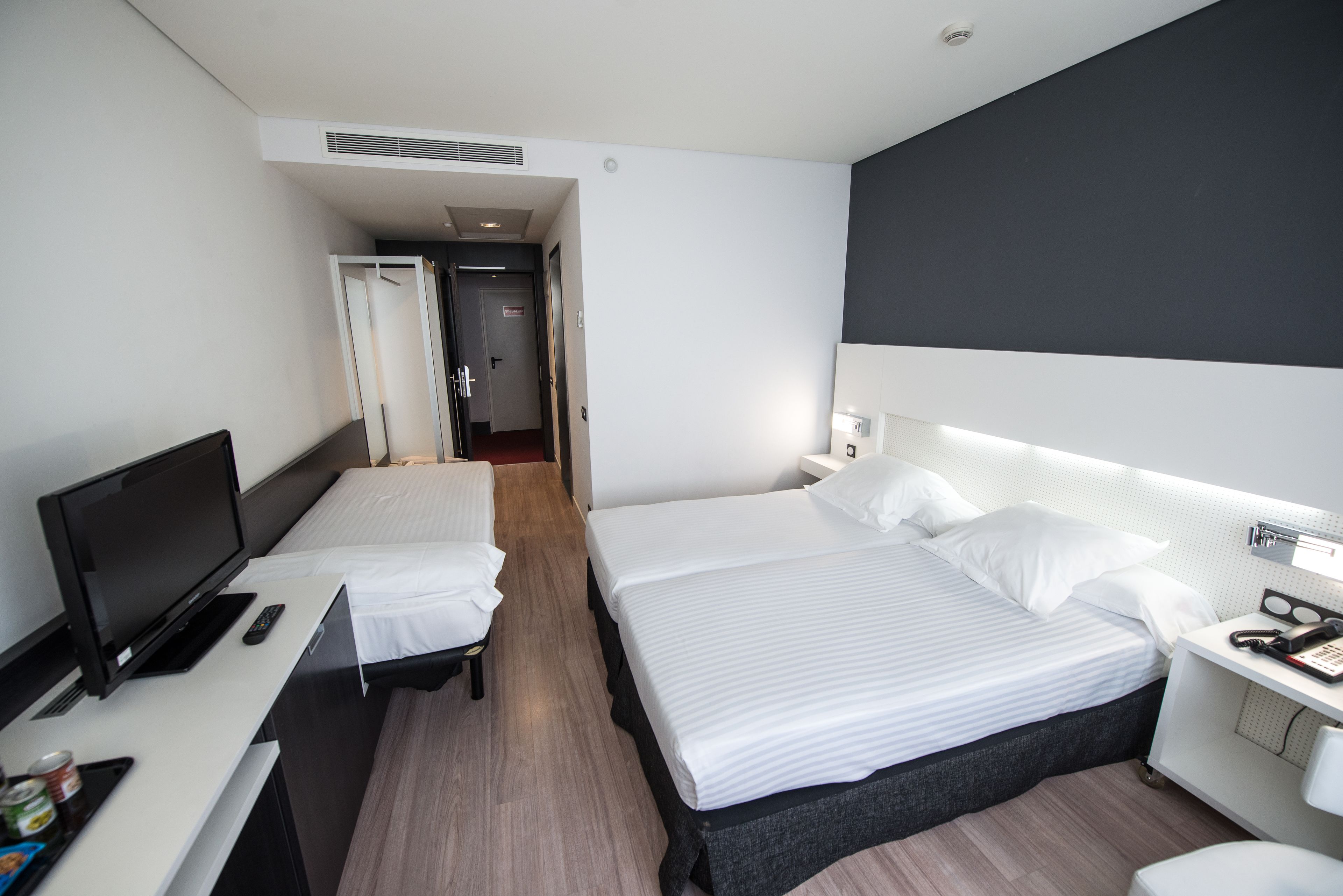 Quarto de hotel com duas camas, uma televisão, uma escrivaninha com computador e uma mesinha de cabeceira com abajur.