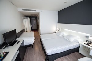 Quarto de hotel com duas camas, uma televisão, uma escrivaninha com computador e uma mesinha de cabeceira com abajur.