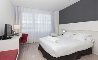 Quarto de hotel com uma cama, uma escrivaninha com computador, uma cadeira e uma televisão.