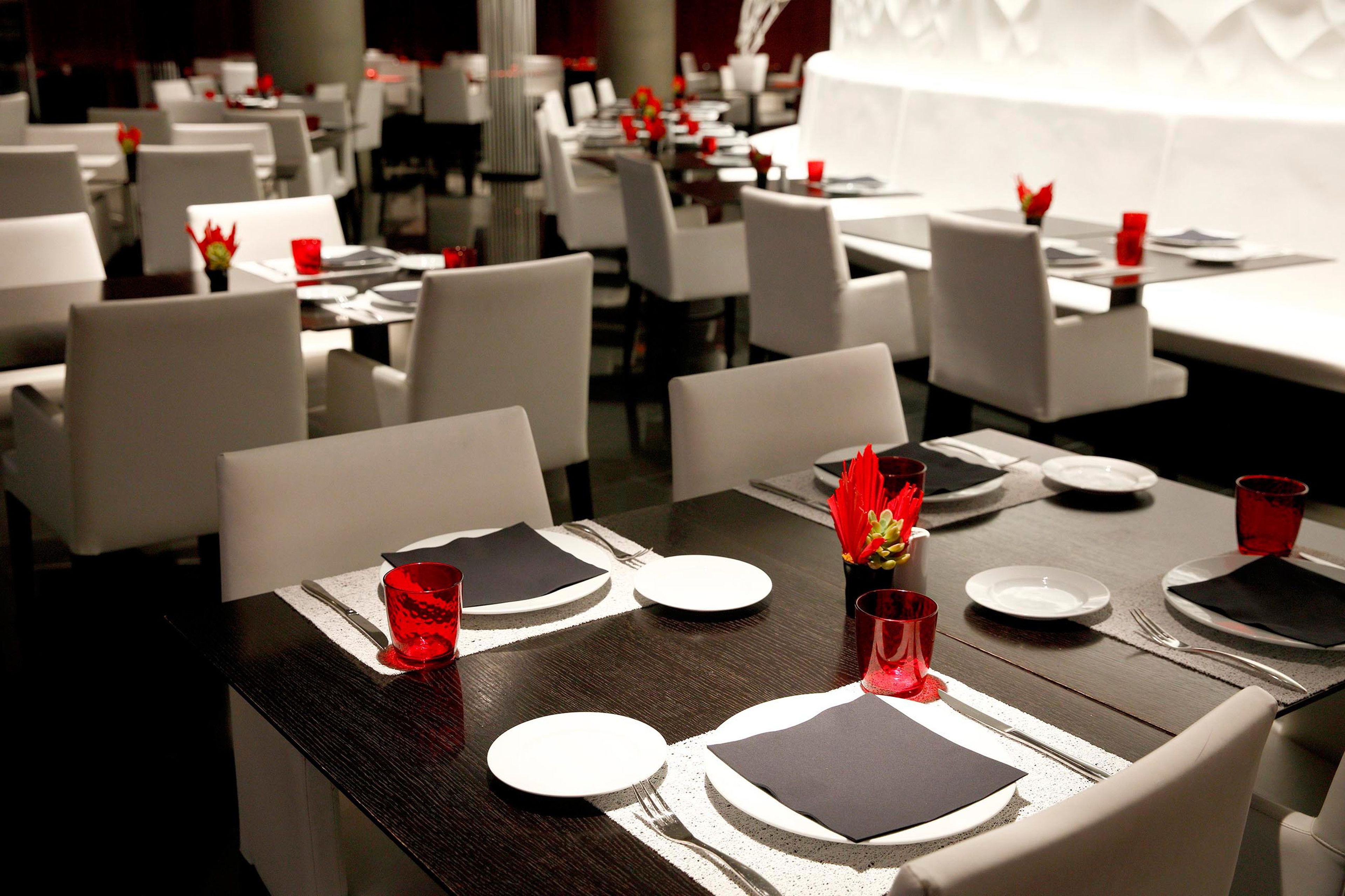 Restaurante com várias mesas preparadas para refeições, cada uma com pratos brancos, guardanapos pretos e copos vermelhos.