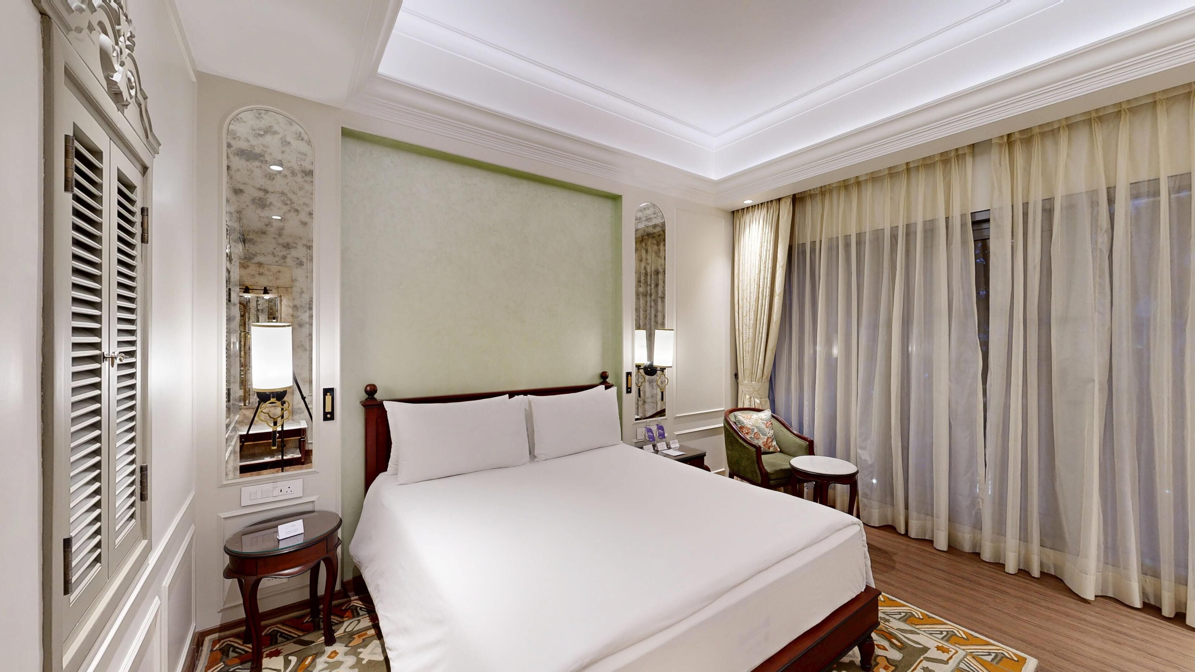 Quarto de hotel com uma cama grande, mesa de cabeceira, uma cadeira, um espelho e uma janela com cortinas.