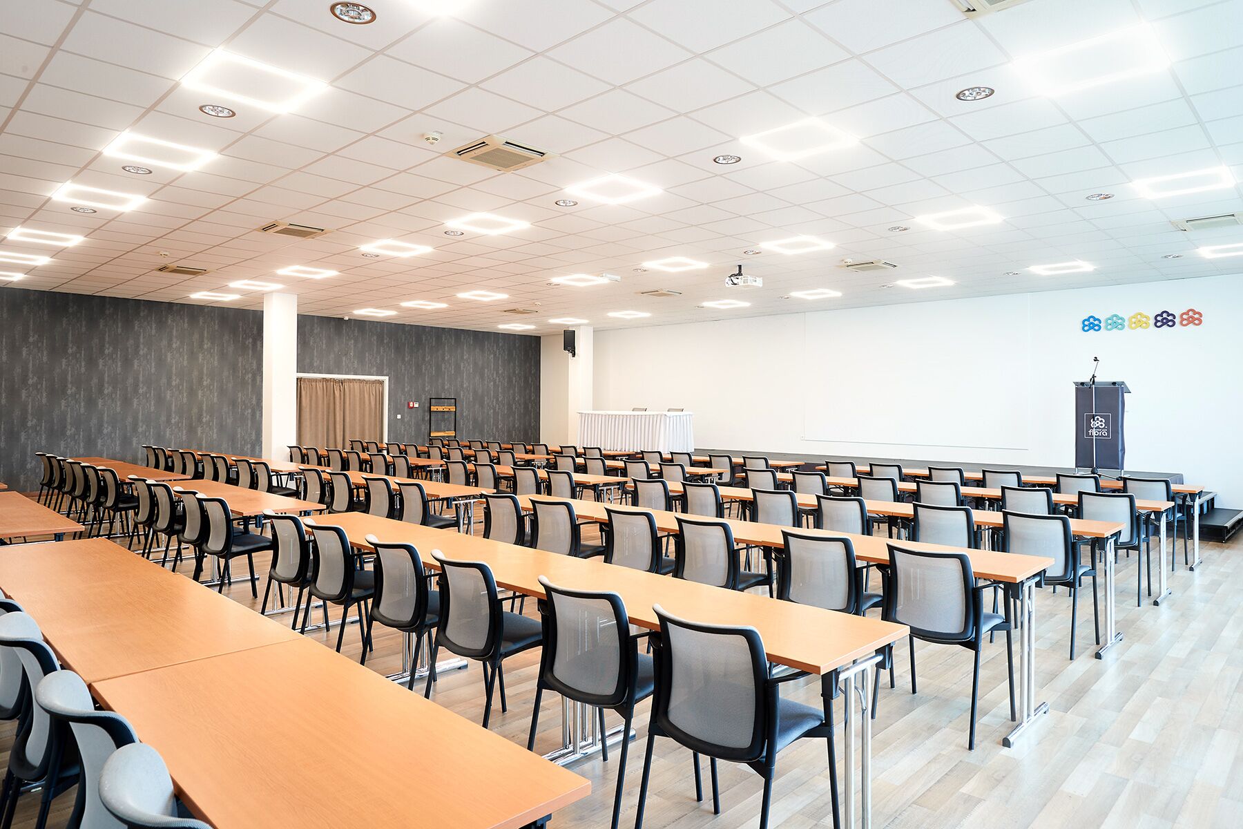 Uma sala de conferência com fileiras de mesas e cadeiras, um pódio e uma tela de projeção.