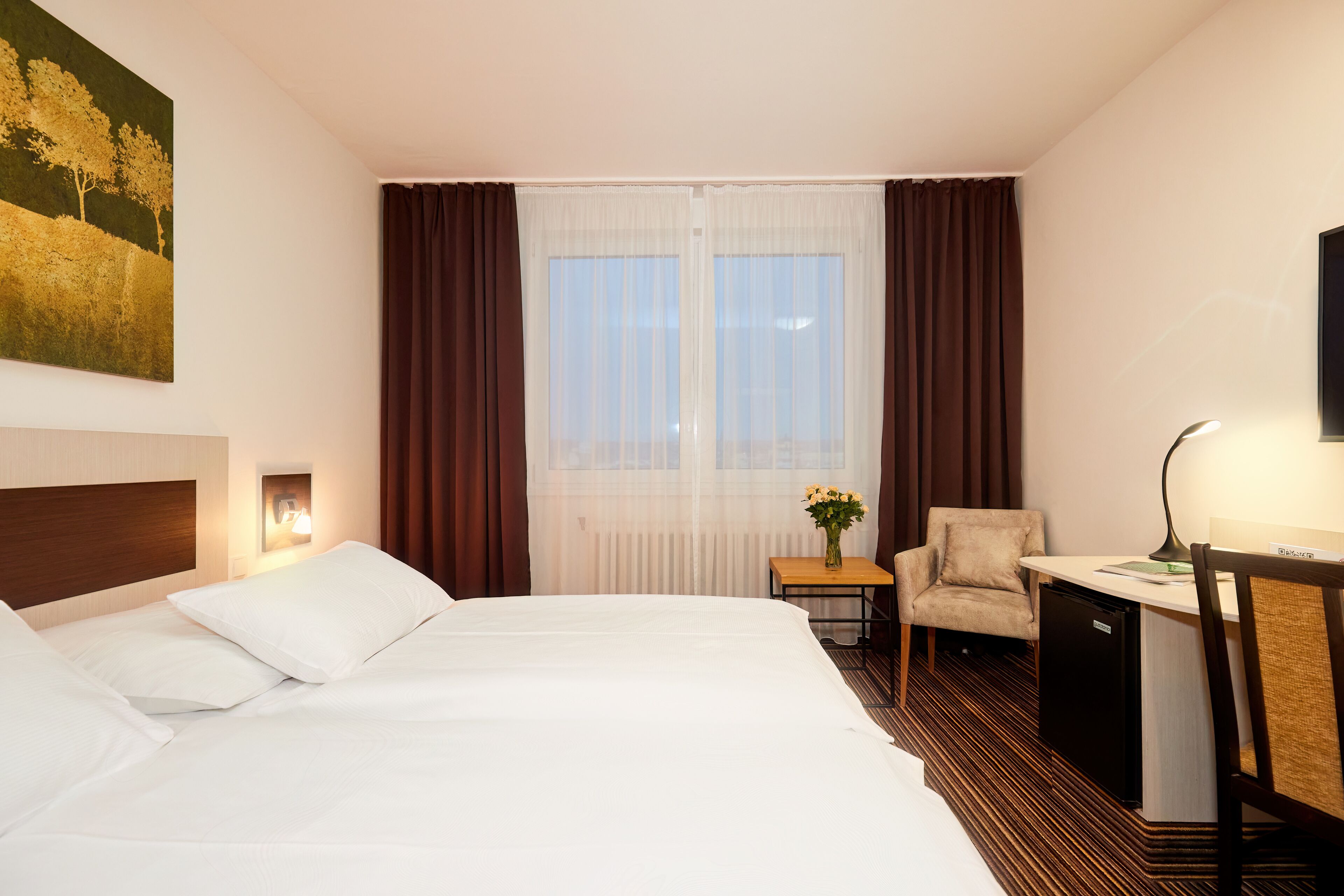 Quarto de hotel com uma cama grande, uma escrivaninha, uma cadeira, uma mesinha de centro com um vaso e uma janela com cortinas.