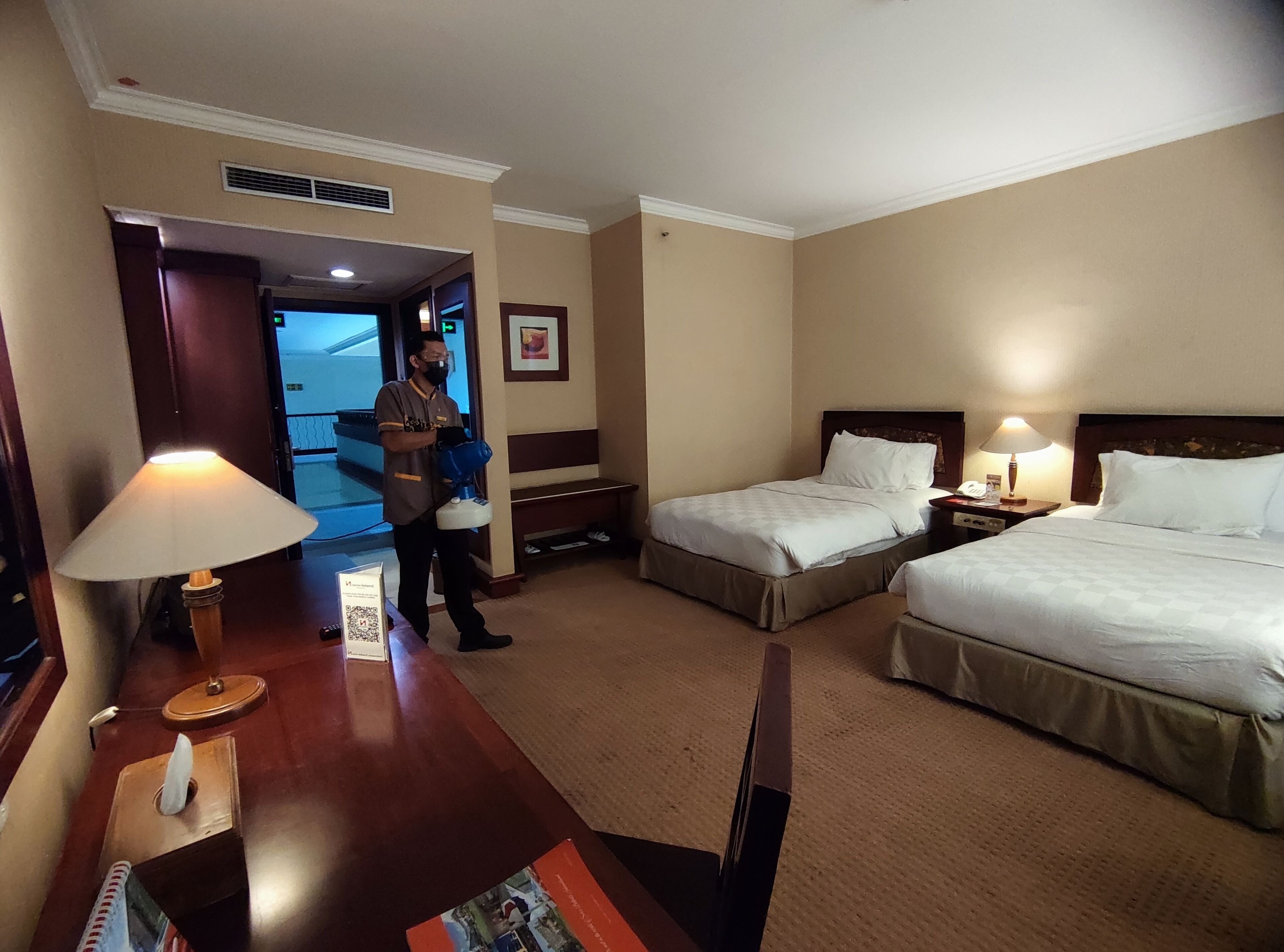 Quarto de hotel com duas camas, uma escrivaninha com luminária, mesa de cabeceira com luminária, um quadro na parede e uma porta que dá para o exterior.