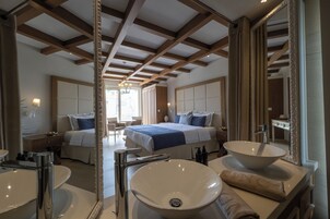Quarto de hotel com uma cama grande, duas mesas de cabeceira, um espelho amplo, uma pia redonda e uma janela.