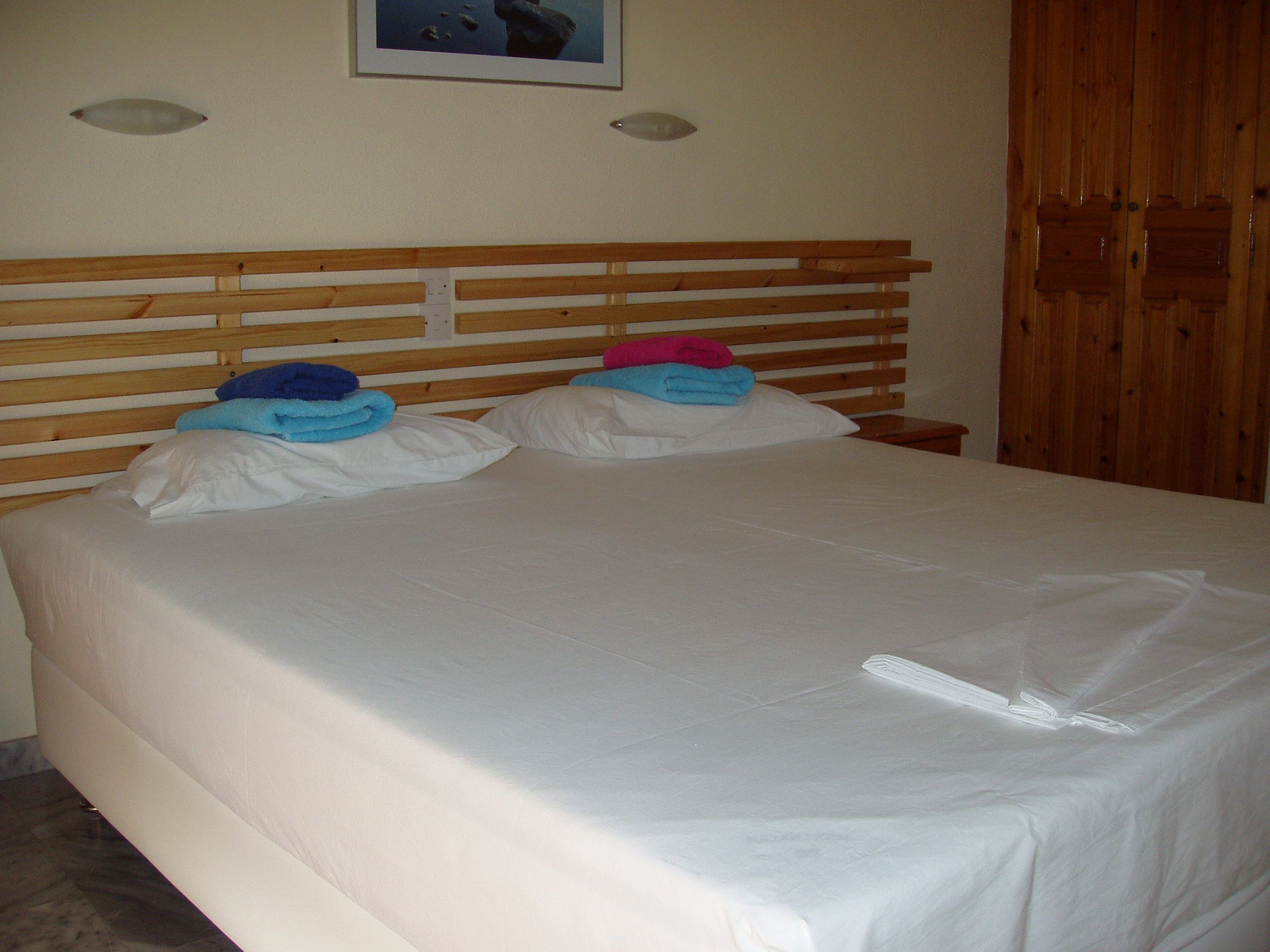 Cama bem arrumada com lençóis brancos e duas almofadas, uma com capa azul e outra com capa rosa.