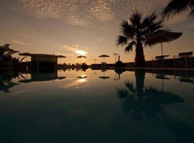 Uma área de piscina tranquila com uma palmeira, guarda-sóis e um edifício refletido na água.