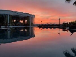 Resort com piscina refletindo o pôr do sol, cadeiras de descanso e um prédio com grandes janelas.