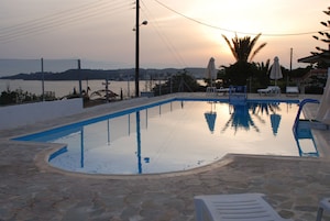 Piscina com deck de pedra, rodeada por espreguiçadeiras e guarda-sóis, com vista para o mar e colinas ao fundo.