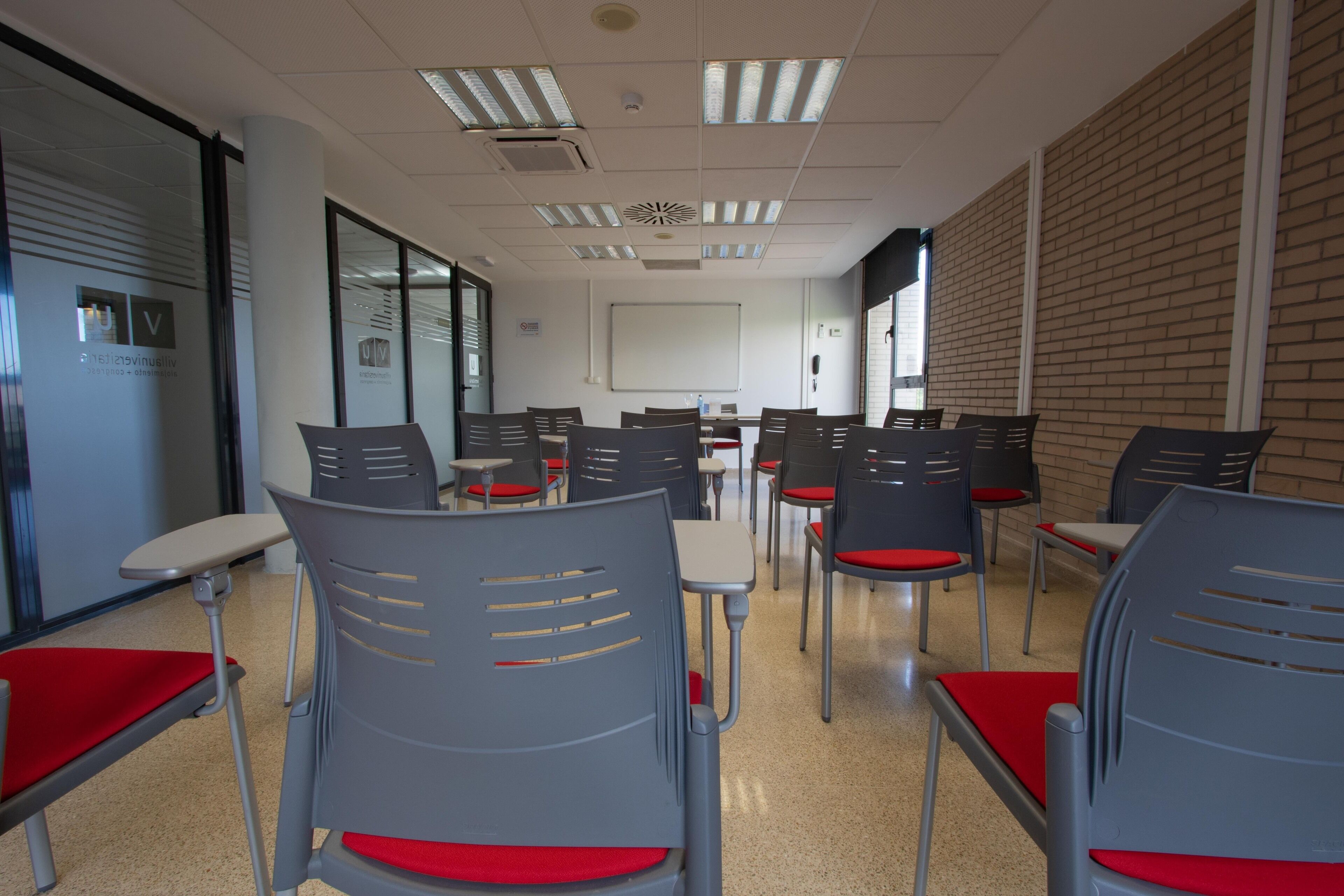 Uma sala de conferências com cadeiras cinzentas e almofadas vermelhas, um quadro branco e uma parede de tijolos.