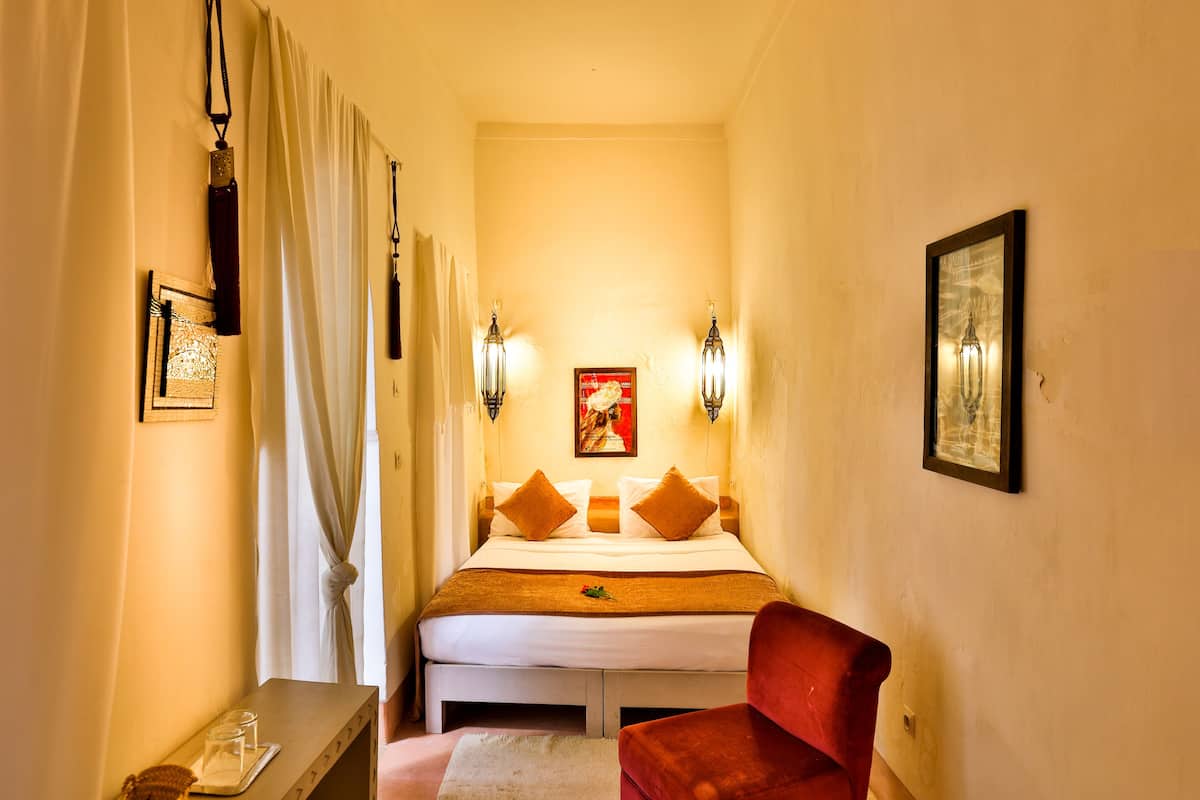 Quarto de hotel estreito, com cama, uma mesinha, uma cadeira e duas luminárias fixadas na parede.