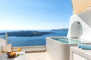 Uma varanda com vista ampla para o mar e ilhas, uma banheira, uma mesa com uma cesta de frutas e um copo de suco de laranja.