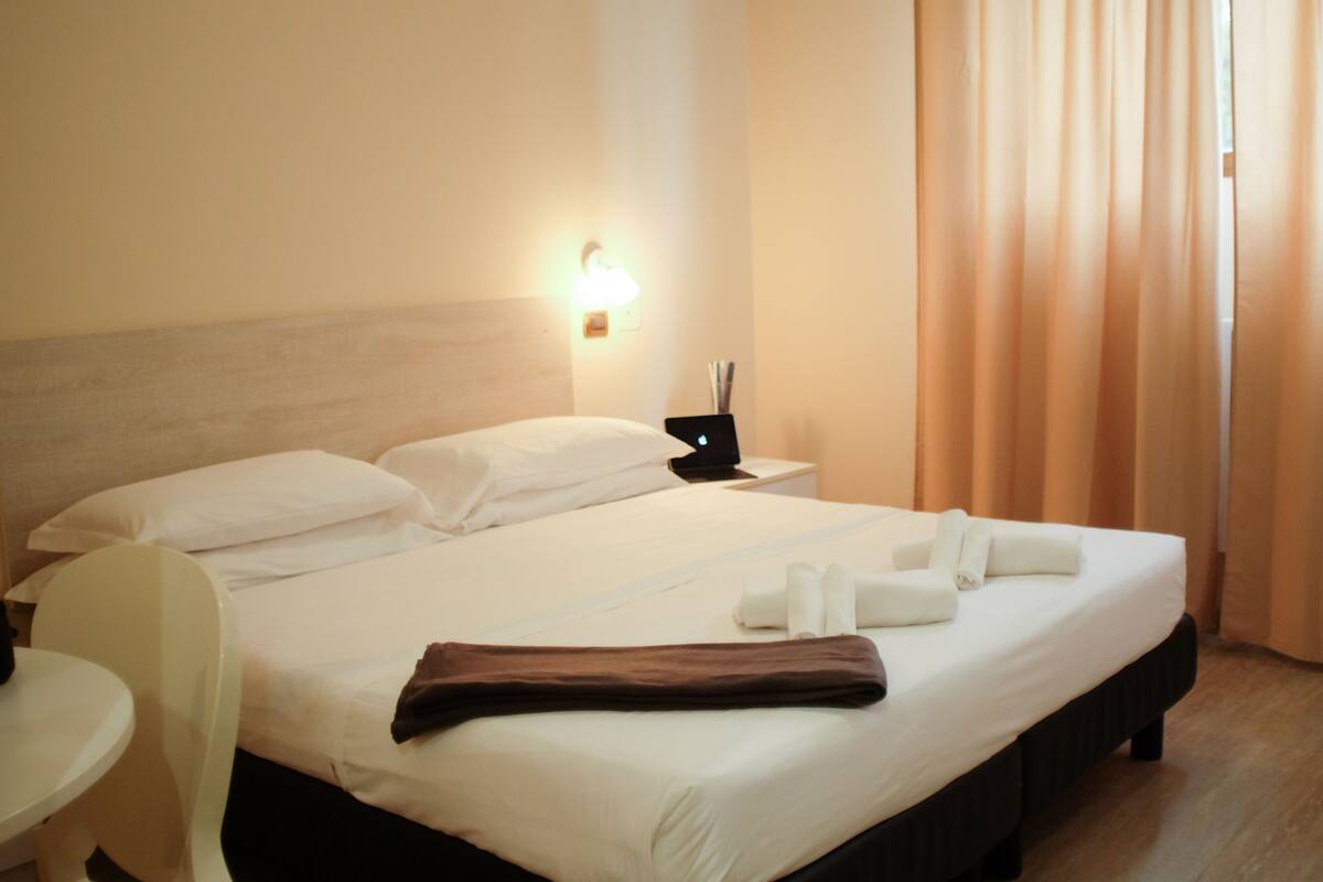 Quarto de hotel com cama, mesa de cabeceira, cadeira e luminária fixada na parede.