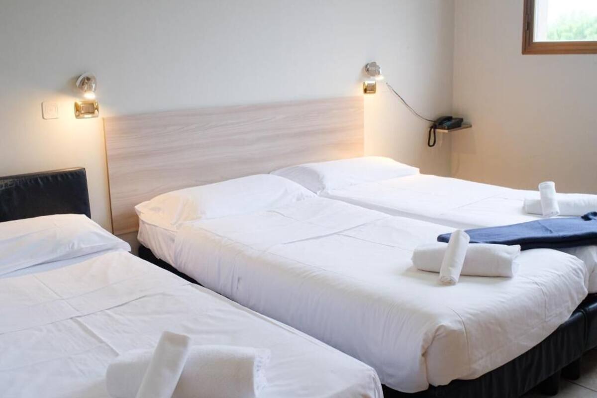 Quarto de hotel com duas camas, cada uma com roupa de cama branca e cabeceira escura. Há luminárias fixadas na parede e um telefone em uma pequena prateleira.