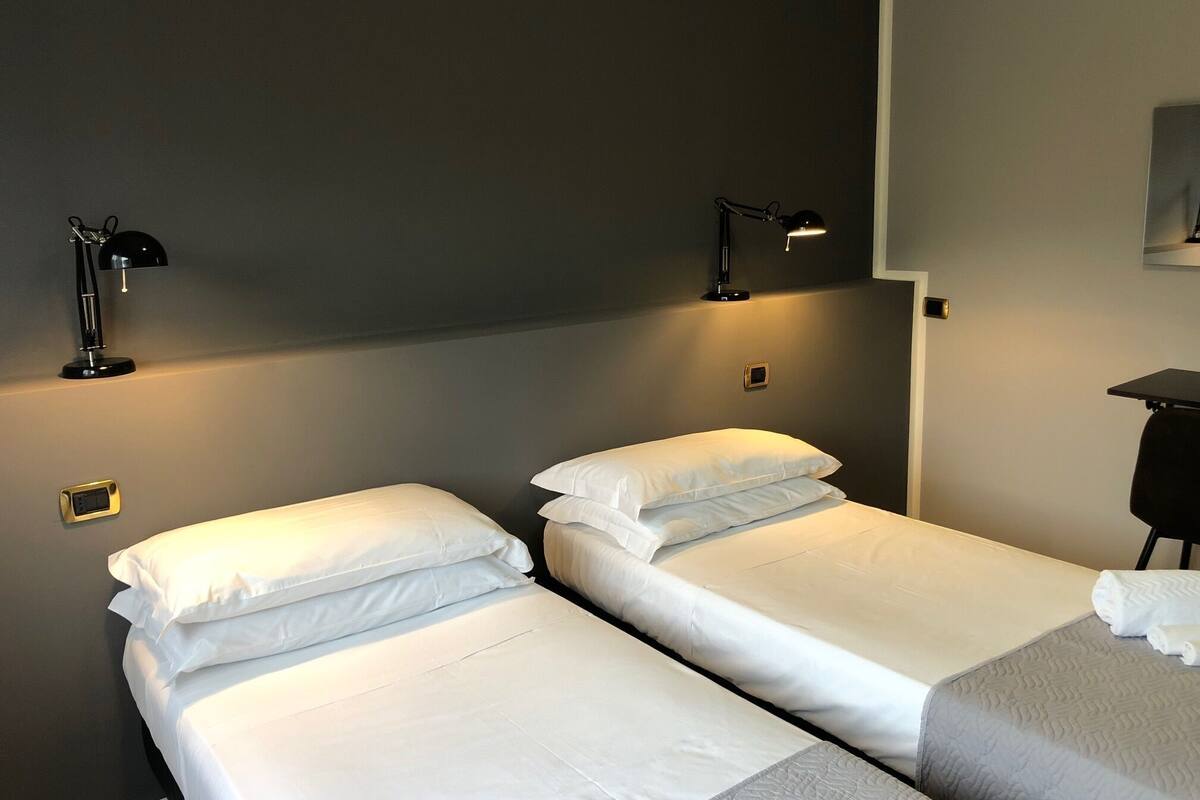 Quarto de hotel com duas camas de solteiro, cada uma com lençóis e almofadas brancos, uma mesa de cabeceira, uma escrivaninha e duas luminárias de leitura fixadas na parede.