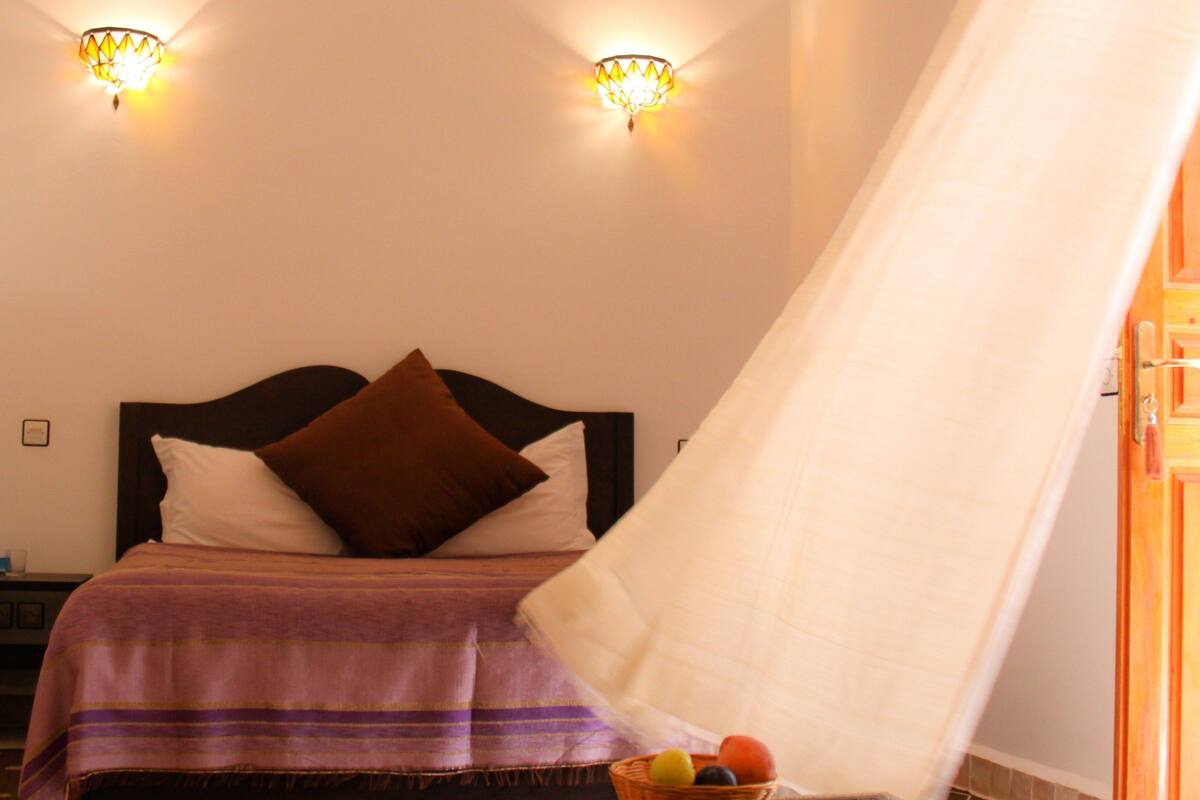 Quarto com cama, travesseiros, cobertor, mesa de cabeceira com cesta de frutas e duas luminárias na parede.