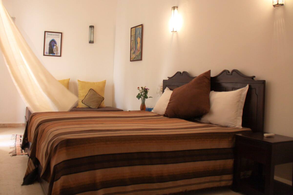 Quarto com cama, almofadas, mesa de cabeceira, luminária na parede e um quadro.