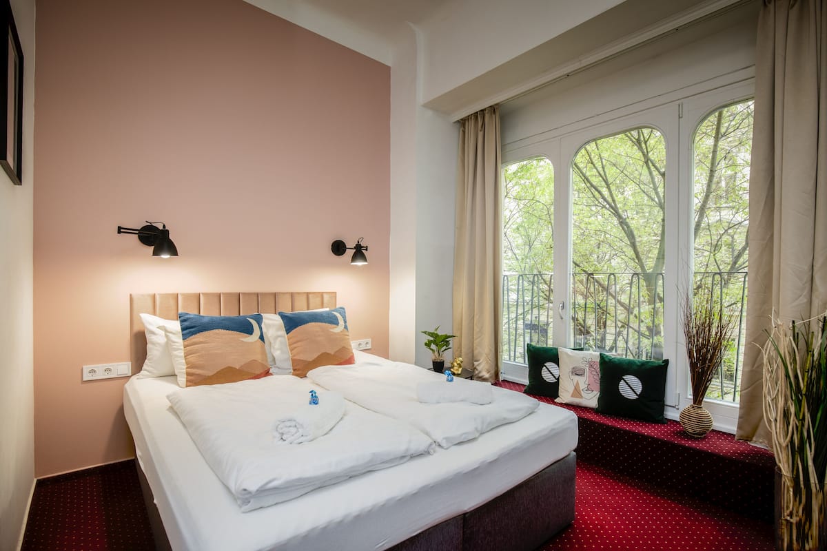 Quarto de hotel com uma cama grande, duas luminárias fixadas na parede, uma janela com cortinas e uma pequena mesa com um vaso.