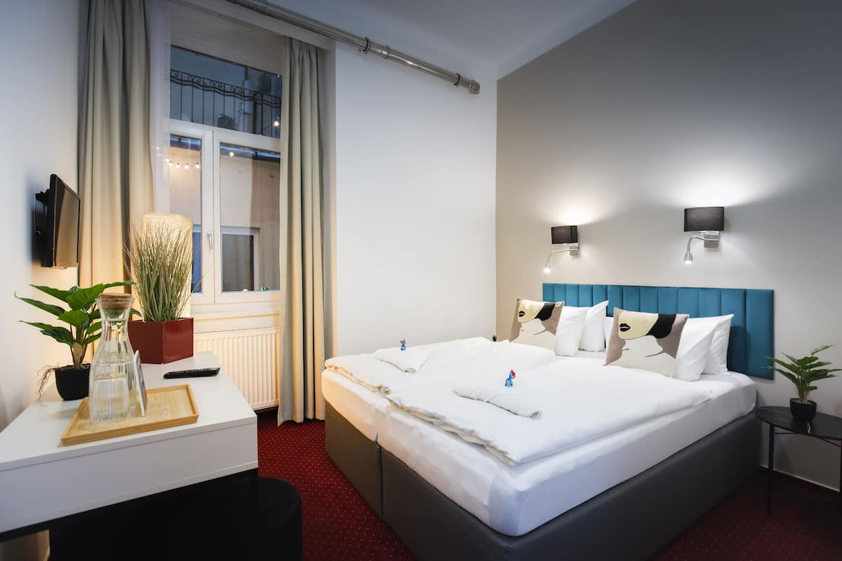 Quarto de hotel com cama, mesas de cabeceira, escrivaninha, planta em vaso, janela com cortinas e luminárias fixadas na parede.